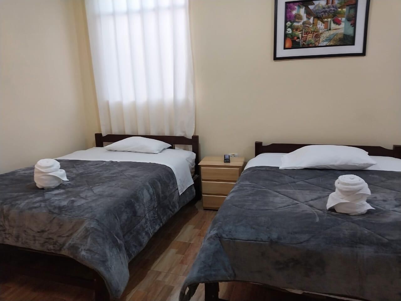 Quarto de hotel com duas camas, cada uma com uma toalha dobrada e um travesseiro branco. Uma pequena mesa de cabeceira de madeira entre as camas possui um telefone. Uma moldura com uma imagem está pendurada na parede acima das camas.