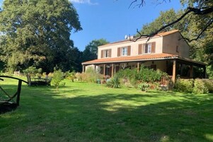 Uma casa de dois andares com varanda, cercada por um gramado bem cuidado e árvores.