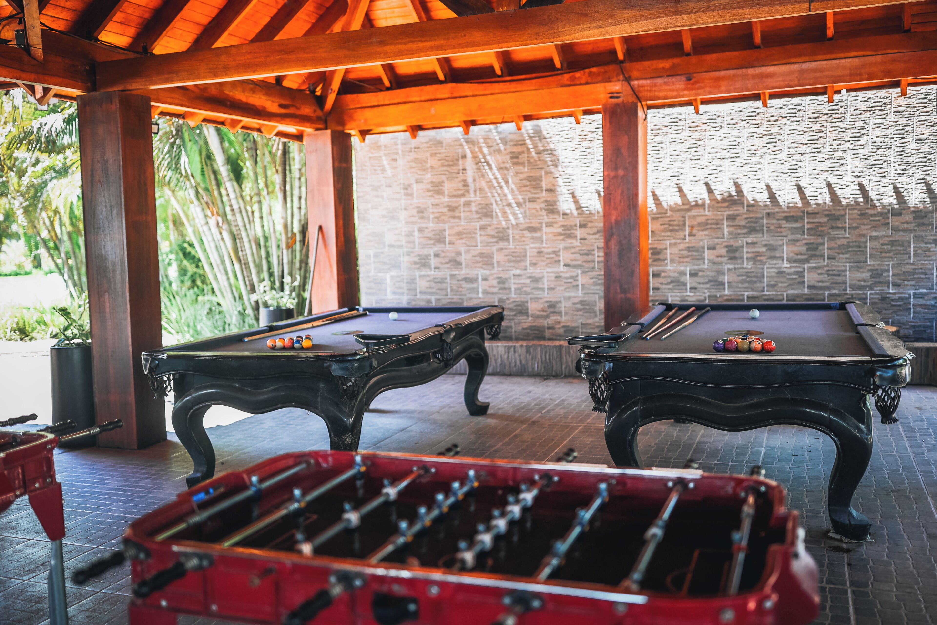 Mesa de bilhar e mesa de futebol de mesa em área externa coberta, com paredes de tijolo e vigas de madeira.