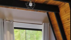 Quarto com uma grande janela, cama com almofadas brancas e estampadas, teto de madeira e uma pequena mesa de cabeceira com abajur.