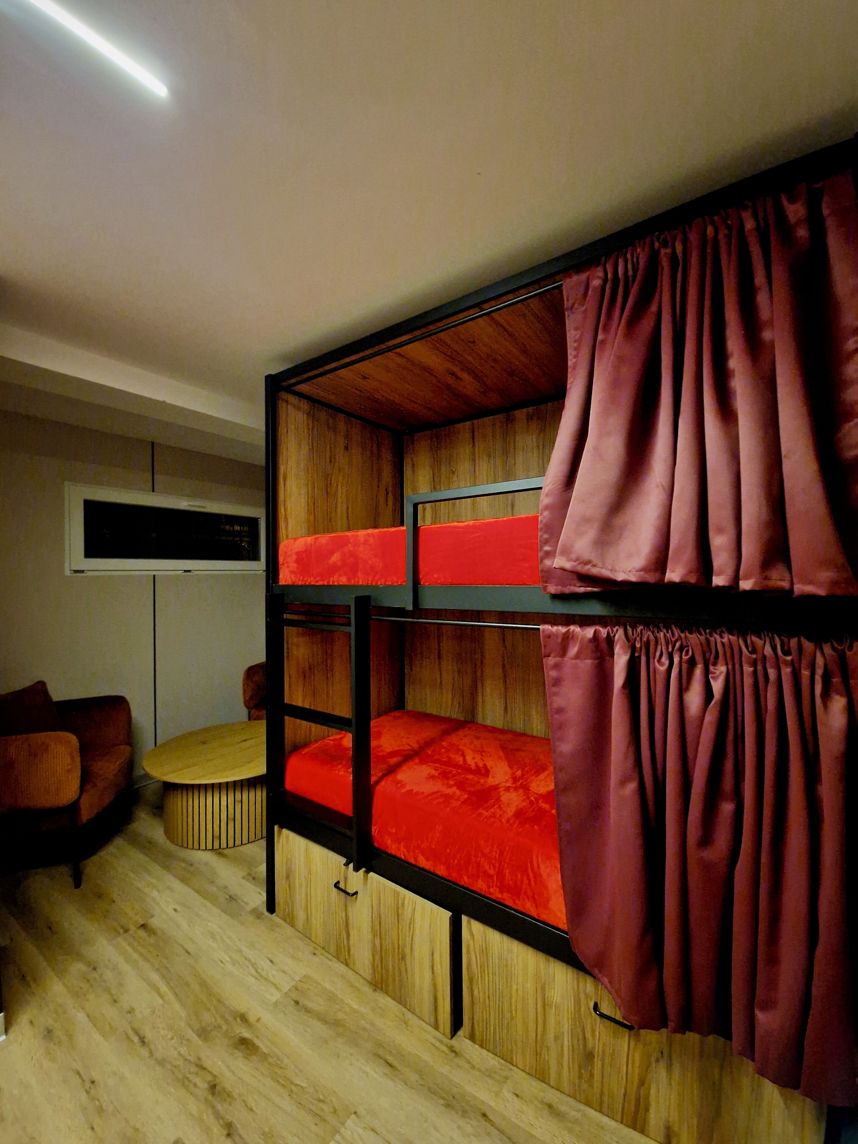 Uma cama beliche com roupa de cama vermelha, chão de madeira, uma pequena mesa redonda e uma cortina vermelha.