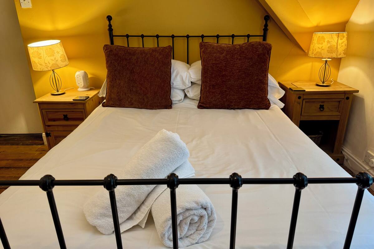 Uma cama bem arrumada com duas almofadas marrons, uma toalha branca e duas mesas de cabeceira com abajures.