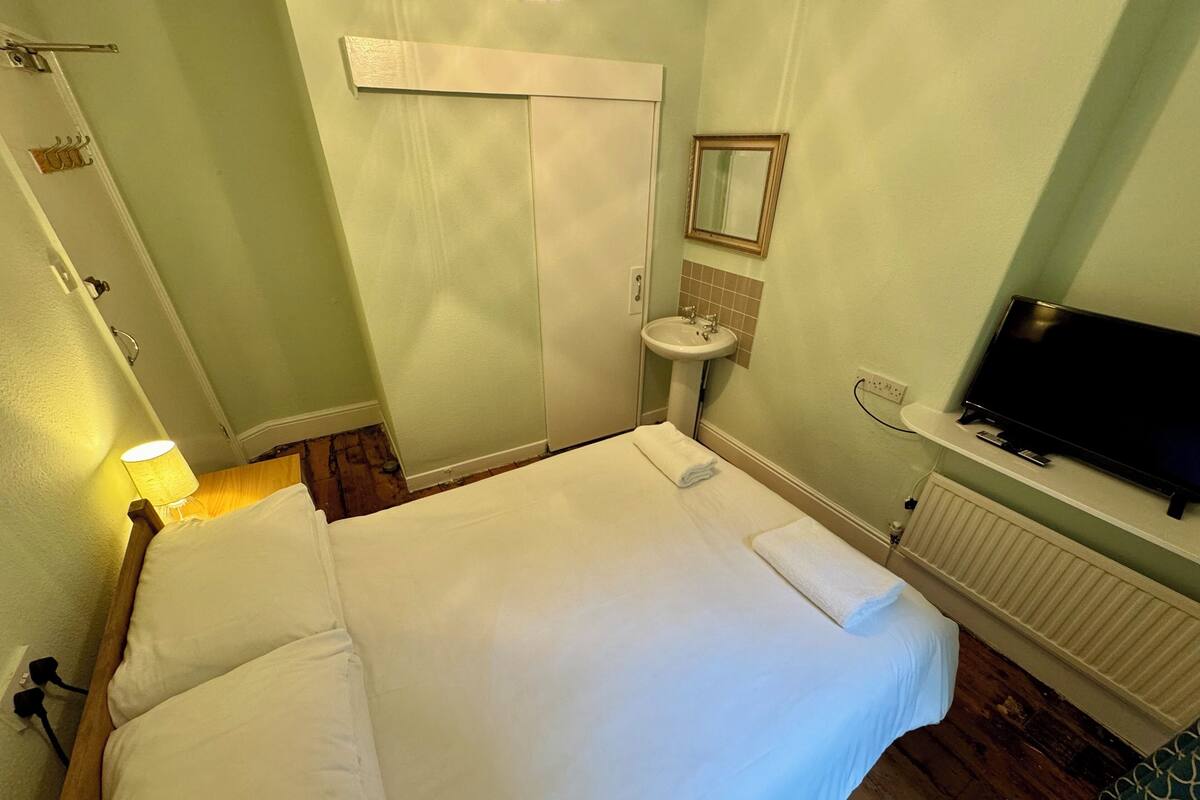 Quarto de hotel com cama, luminária de cabeceira, pia, espelho e televisão.