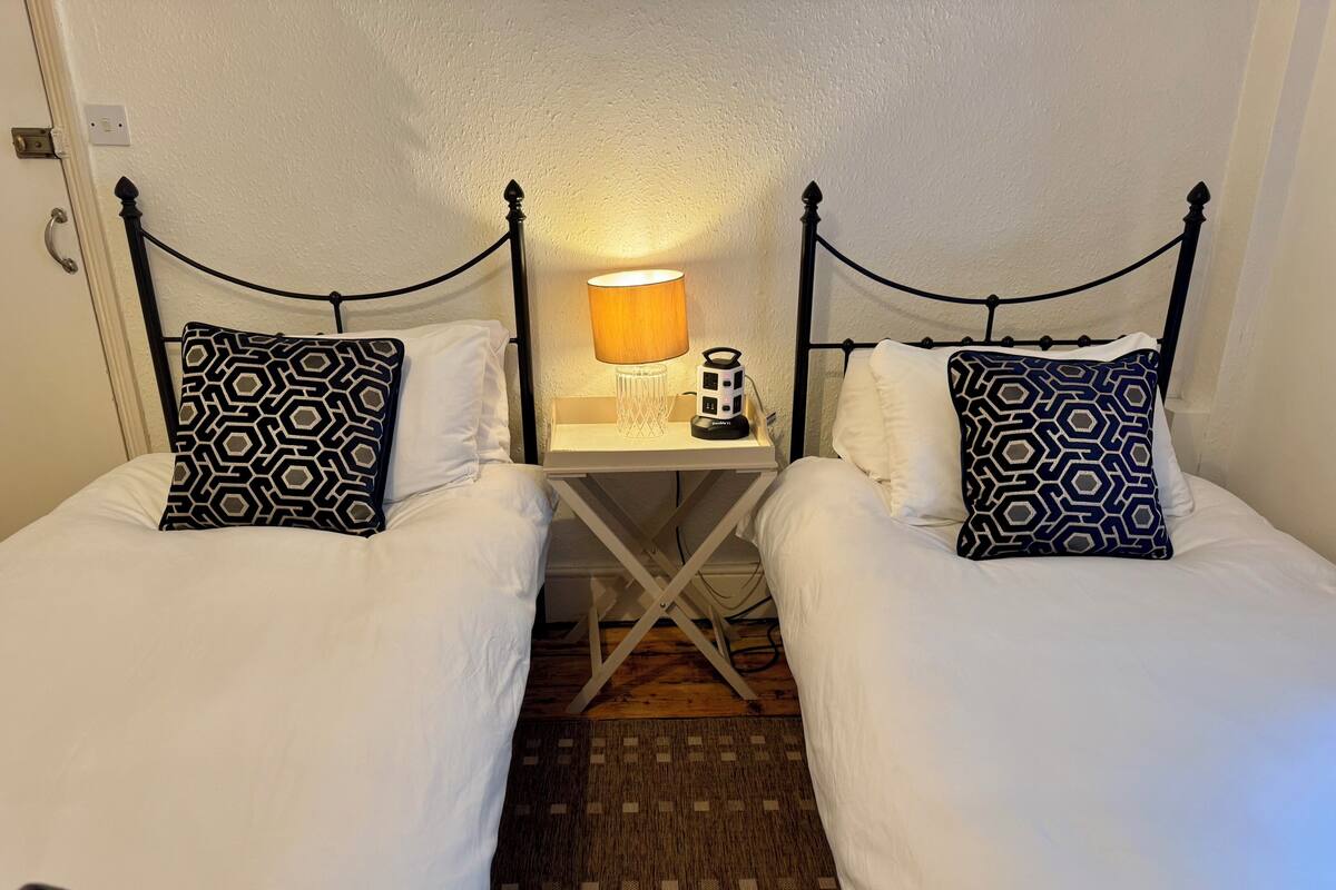 Quarto de hotel com duas camas de solteiro, cada uma com almofada estampada e mesa de cabeceira com abajur e pequeno dispositivo eletrônico.