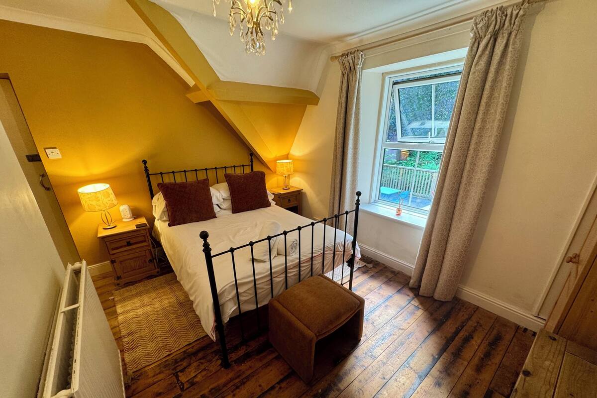 Quarto com cama, luminárias de cabeceira, janela com cortinas, radiador e piso de madeira.