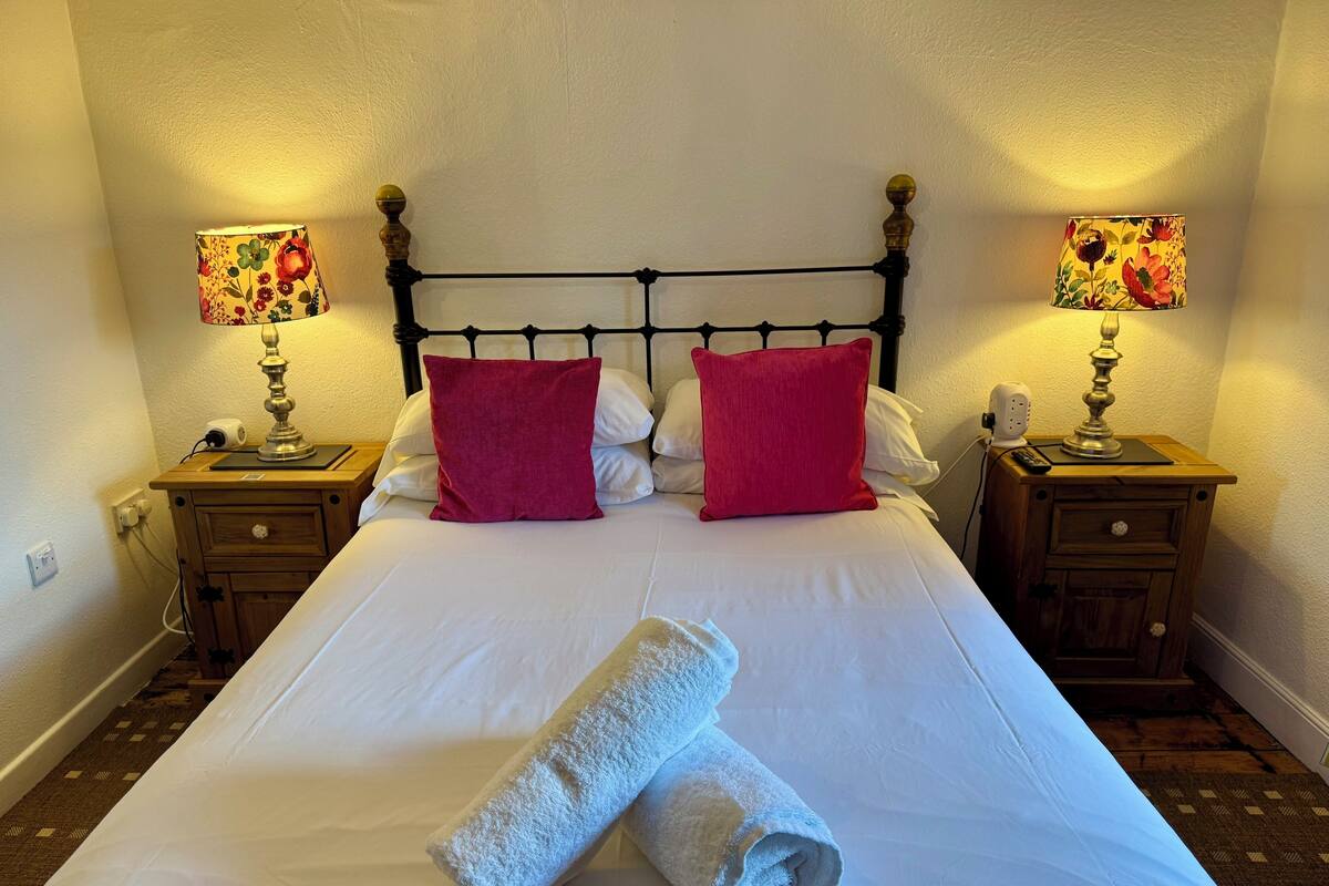 Cama bem arrumada com lençóis brancos, dois travesseiros vermelhos e uma toalha dobrada. Há duas mesas de cabeceira com luminárias, cada uma com um abajur floral. O quarto tem um design simples e limpo, com cabeceira de madeira e parede lisa.