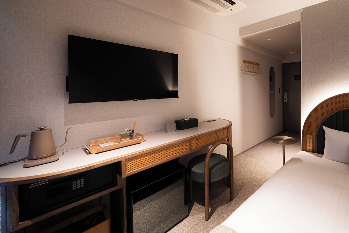 Quarto de hotel com televisão de tela plana na parede, escrivaninha com chaleira, mesa pequena, cama com travesseiro estampado e cabeceira com ripas verticais.