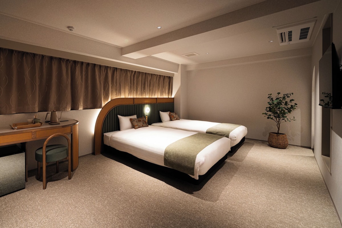 Quarto de hotel com escrivaninha, duas camas, um espelho grande e um vaso de planta.