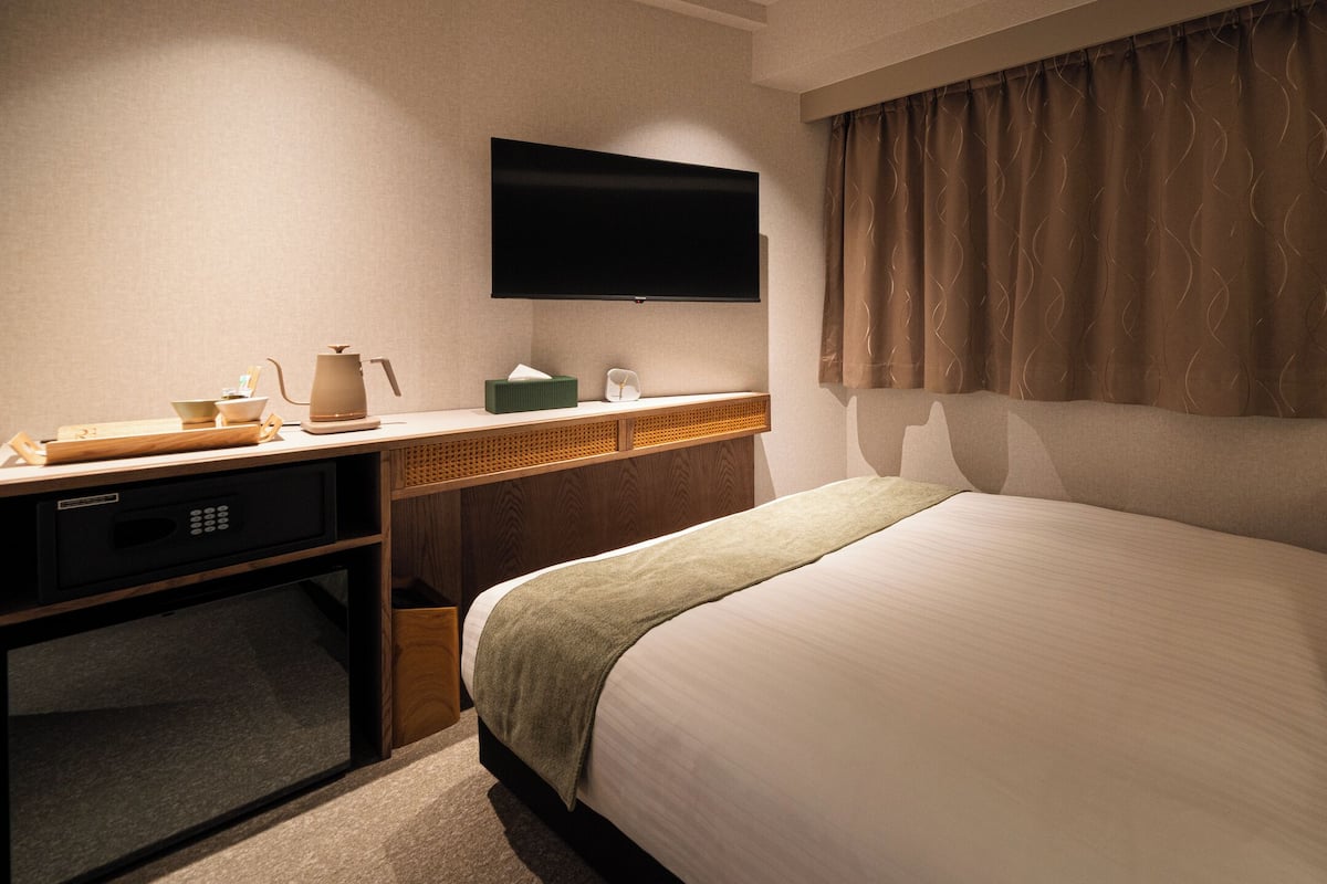 Quarto de hotel com cama, escrivaninha com cafeteira, televisão e janela com cortinas.