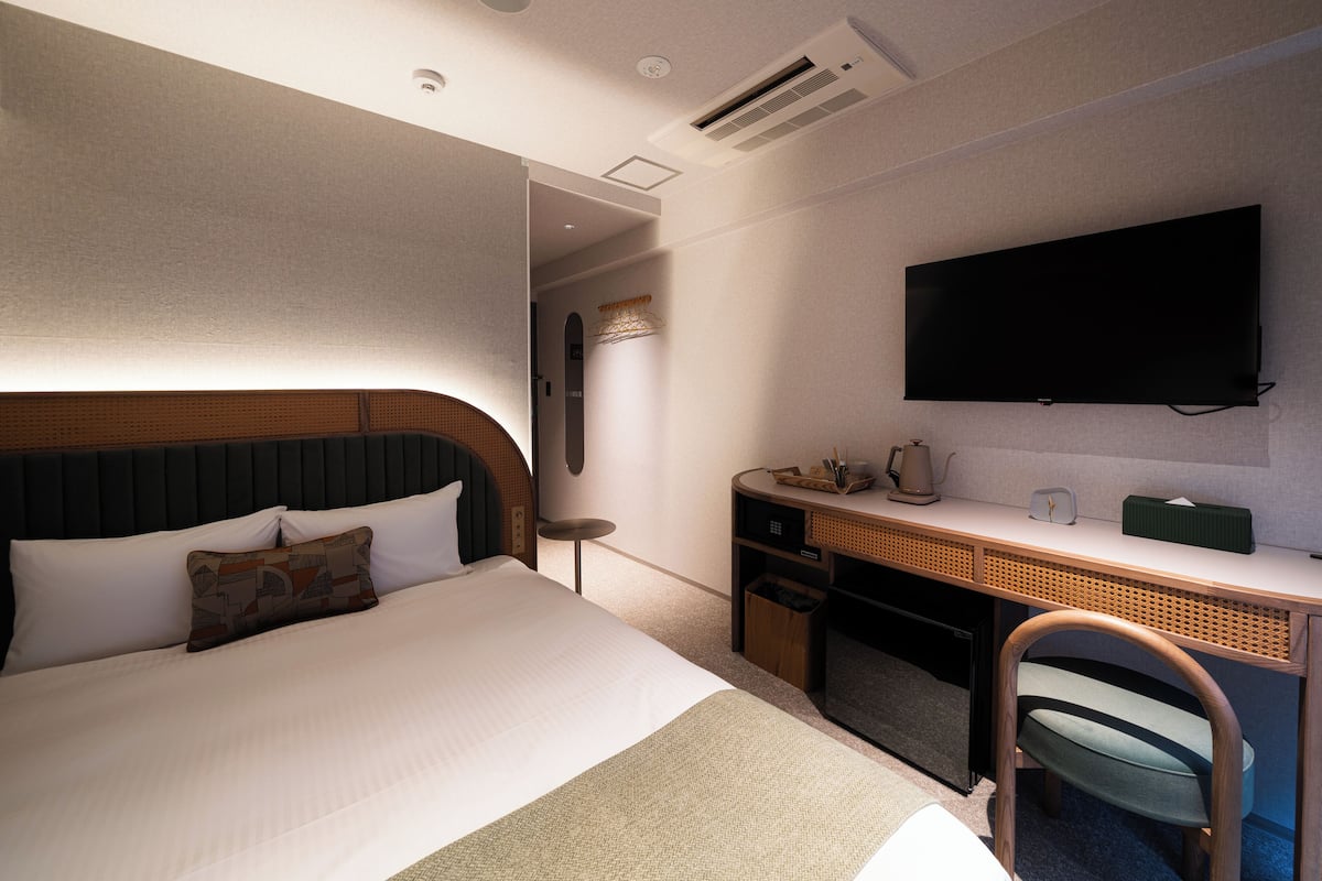 Quarto de hotel com cama, escrivaninha, cadeira, televisão e lixeira.