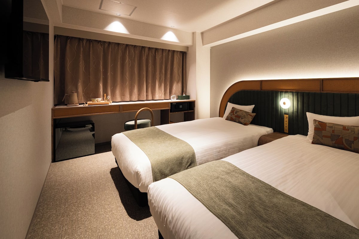 Quarto de hotel com duas camas, uma escrivaninha, uma cadeira, televisão e uma janela com cortinas.