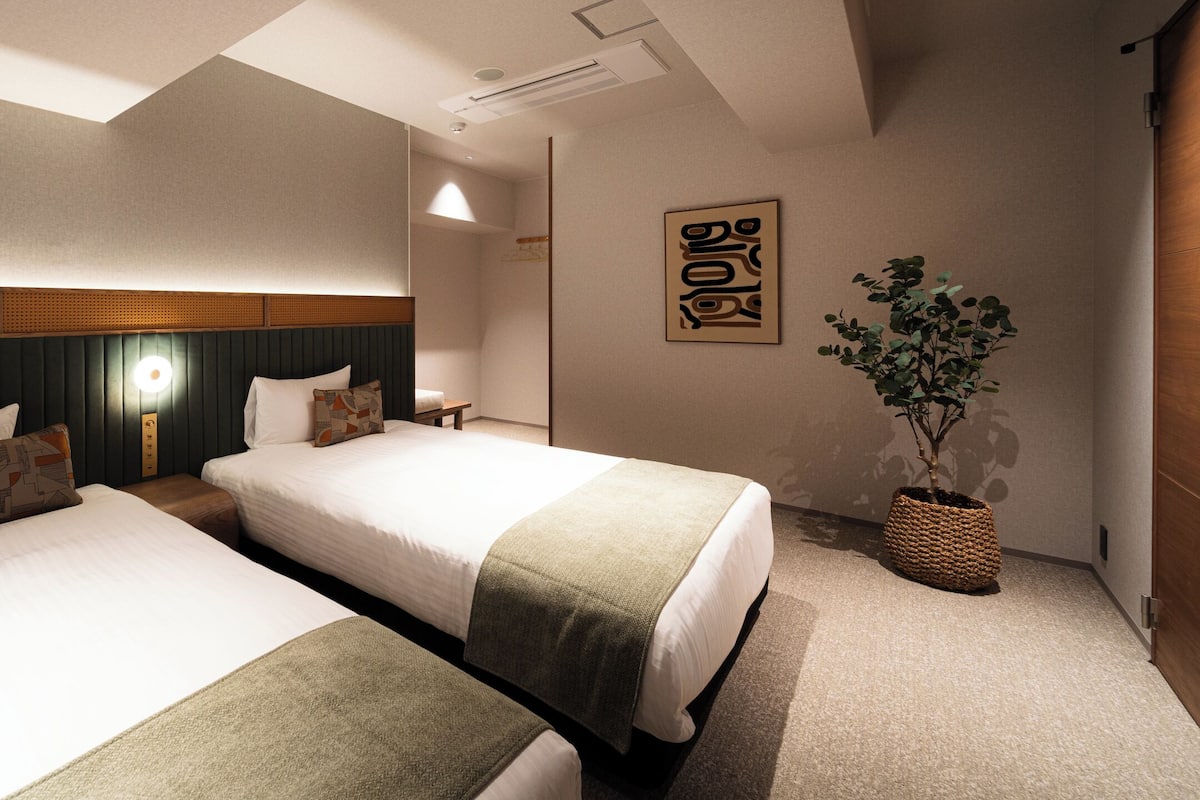 Quarto de hotel com duas camas, uma cabeceira de madeira, uma obra de arte na parede e uma planta em vaso.