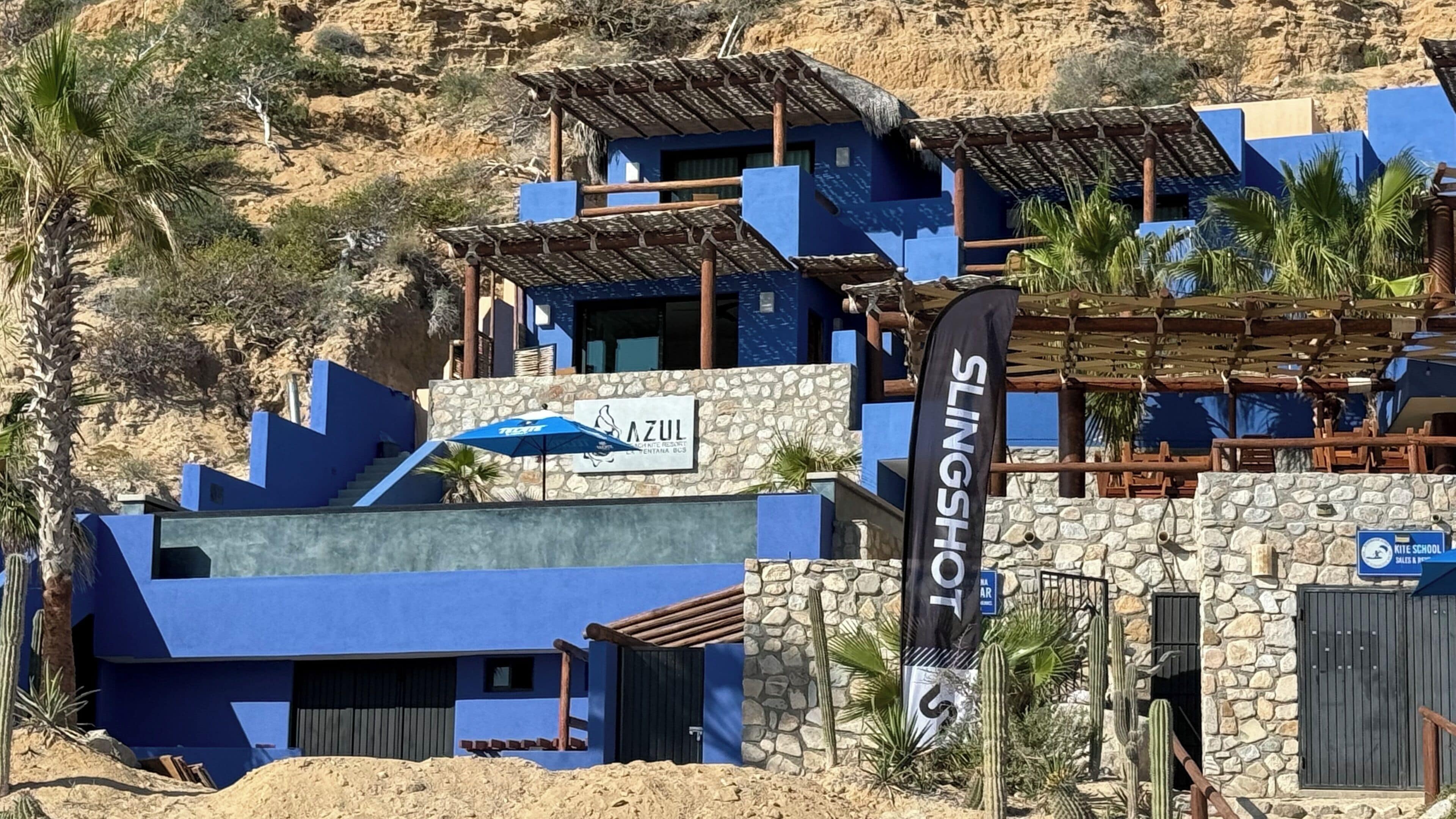 Um edifício com fachada de pedra e detalhes azuis, um letreiro com 'AZUL' e uma bandeira com 'KITE SCHOOL SALES & SERVICE'.