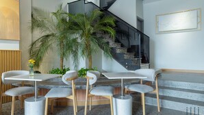 Um interior moderno com área de jantar, mesa branca e cadeiras cinzas, escada com guarda-corrimão de vidro e uma planta grande.