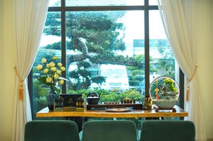 Quarto com vista para árvores e uma van branca através de uma grande janela, uma mesa com um vaso de flores e um objeto decorativo sobre a mesa.