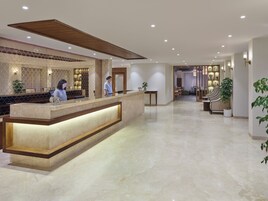 Uma área de recepção de hotel com balcão de mármore, mesa de atendimento e espaço de estar com cadeiras e mesa.