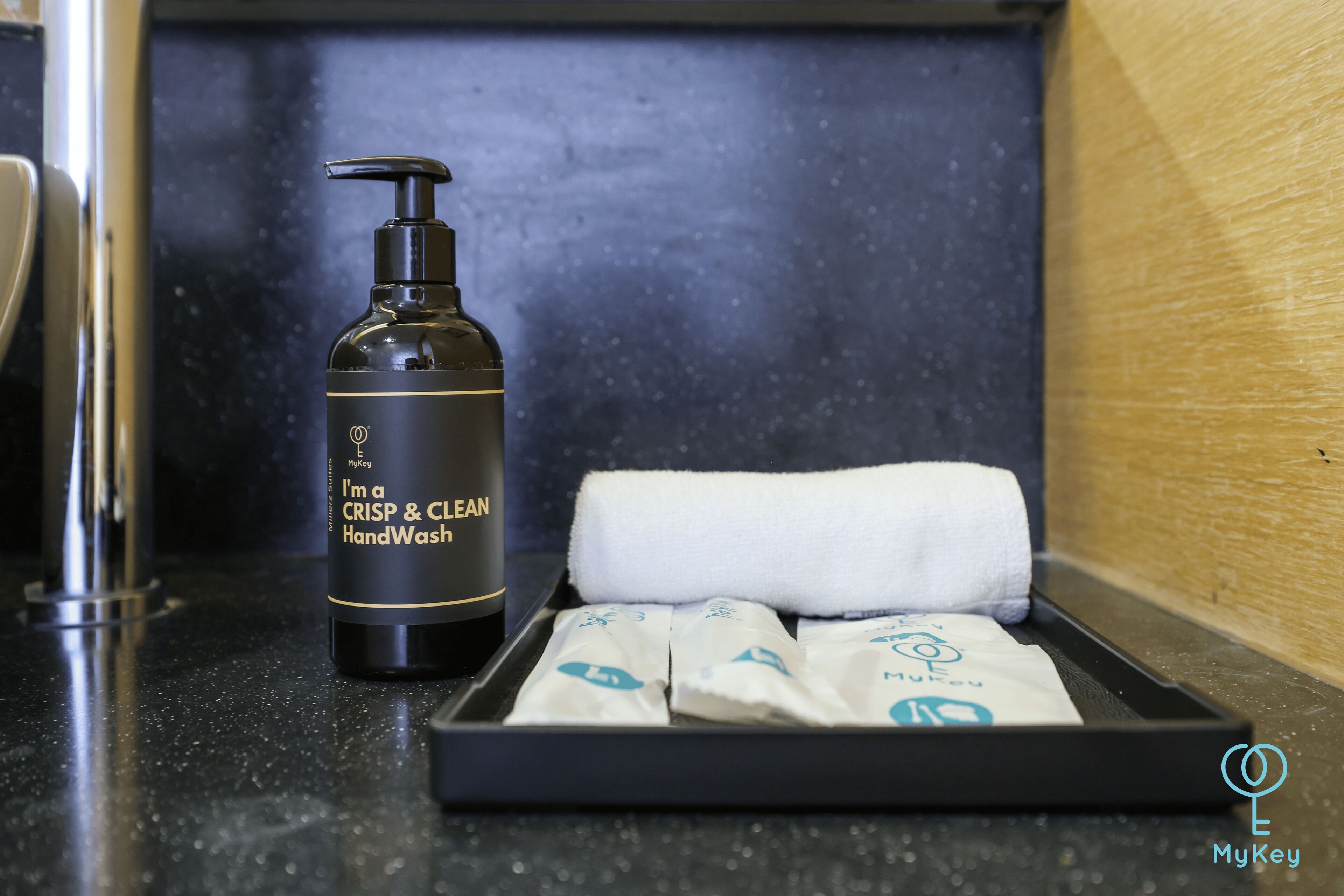 Um dispensador de sabonete líquido, uma toalha branca e quatro embalagens de sabonete em um prato preto em um banheiro.