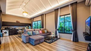 Sala moderna com sofá seccional, mesa de centro e televisão. Há uma área de cozinha com máquina de lavar e mesa de jantar. O quarto possui janelas amplas com cortinas e um lustre.