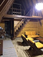 Interior de uma cabana de madeira com quarto no mezanino, área de jantar e escada.
