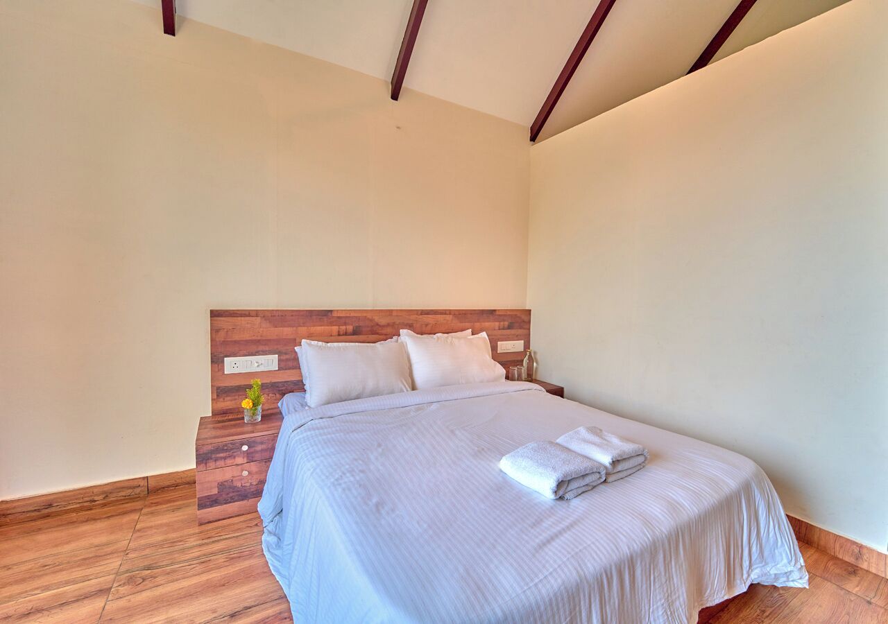 Uma cama bem arrumada com lençóis brancos e uma toalha dobrada, uma cabeceira de madeira e uma pequena mesa de cabeceira com um vaso de flores.