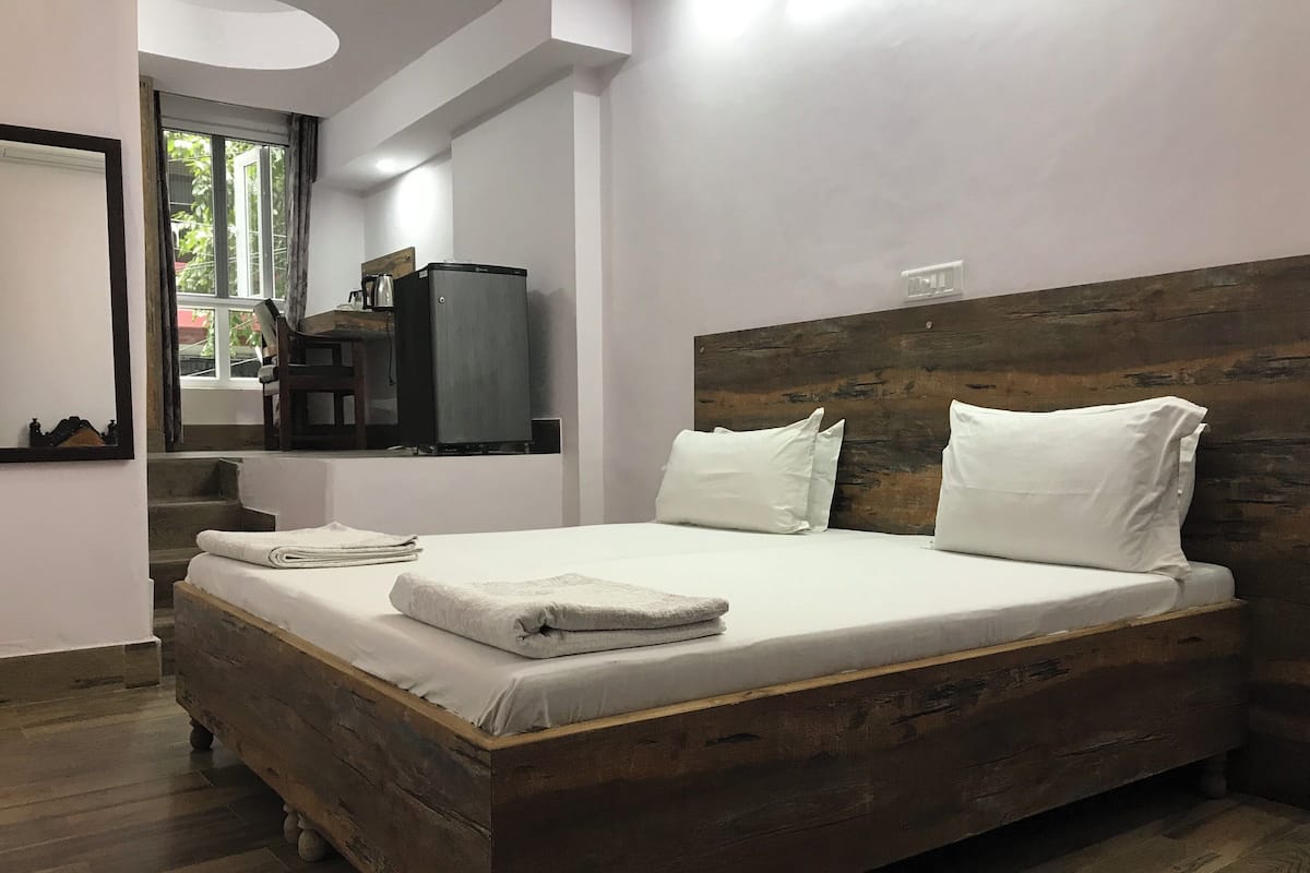 Quarto com cama de madeira, roupa de cama branca e pequena área de cozinha compacta com cafeteira.