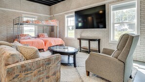 Uma sala de estar espaçosa com teto alto, uma grande TV de tela plana na parede, um sofá confortável, uma mesa de centro redonda e uma cama beliche com roupa de cama laranja.