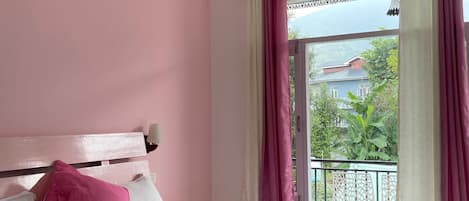 Quarto com cama, ventilador de teto, janela com cortinas e vista para o verde lá fora.