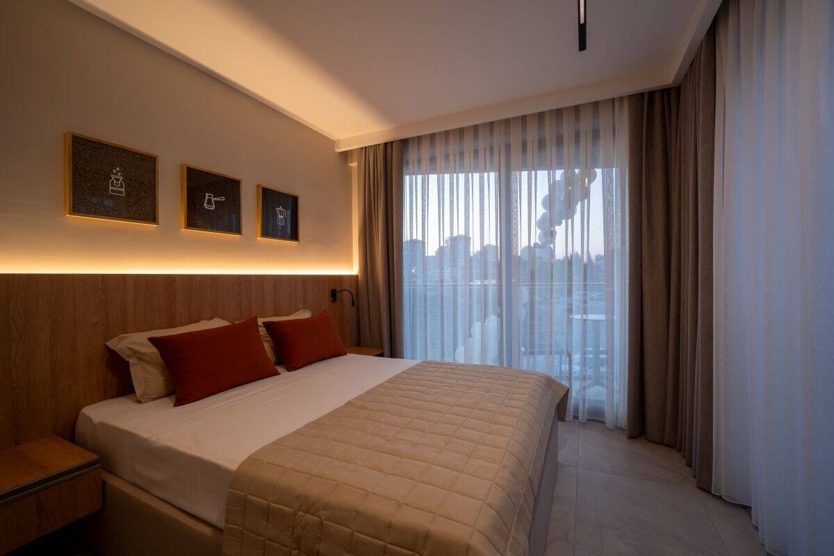 Quarto de hotel com uma cama grande, mesa de cabeceira e vista para a cidade através de cortinas translúcidas.