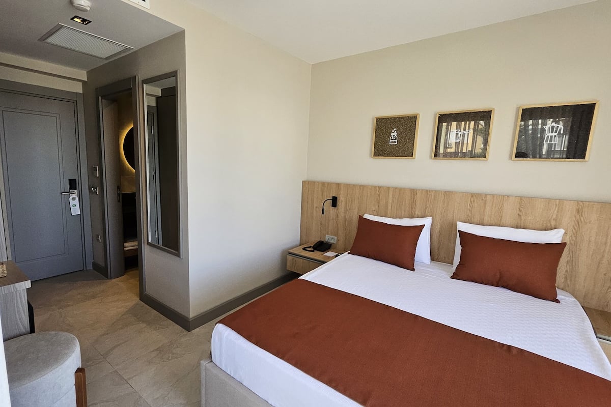 Quarto de hotel com uma cama grande, mesa de cabeceira, escrivaninha e uma cadeira. Há duas imagens emolduradas na parede.