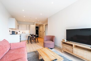 Uma sala de estar moderna com televisão de tela plana, um sofá rosa, uma mesa de centro de madeira e uma cozinha ao fundo.