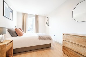 Quarto moderno com uma cama grande, cômoda de madeira e vista para a cidade pela janela.