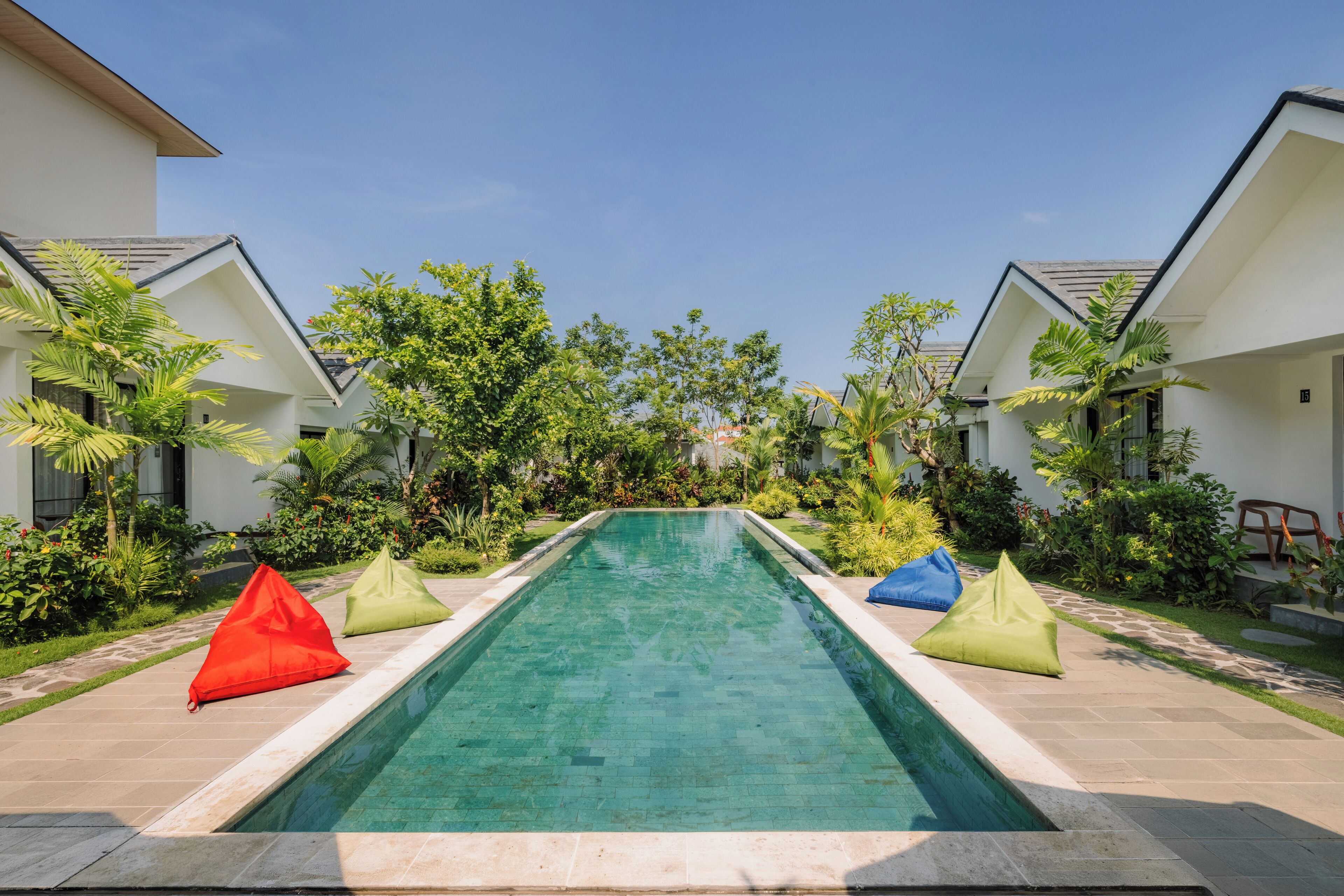 Uma piscina com superfície de água azul cristalina, rodeada por vegetação exuberante e dois edifícios brancos com telhados inclinados.