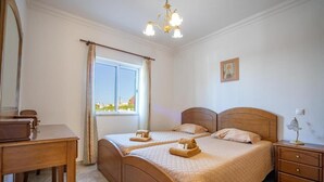 Quarto com duas camas, uma escrivaninha, uma cadeira, mesa de cabeceira e janela com cortinas.