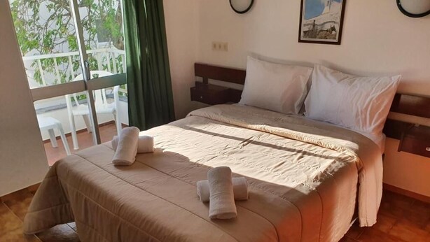 Cama bem arrumada com colcha bege e travesseiros, cabeceira de madeira e vista para o exterior por uma janela grande.
