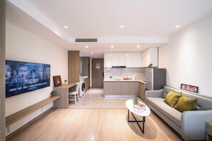 Sala de estar moderna com televisão de tela plana, sofá, mesa de centro e área de cozinha ao fundo.