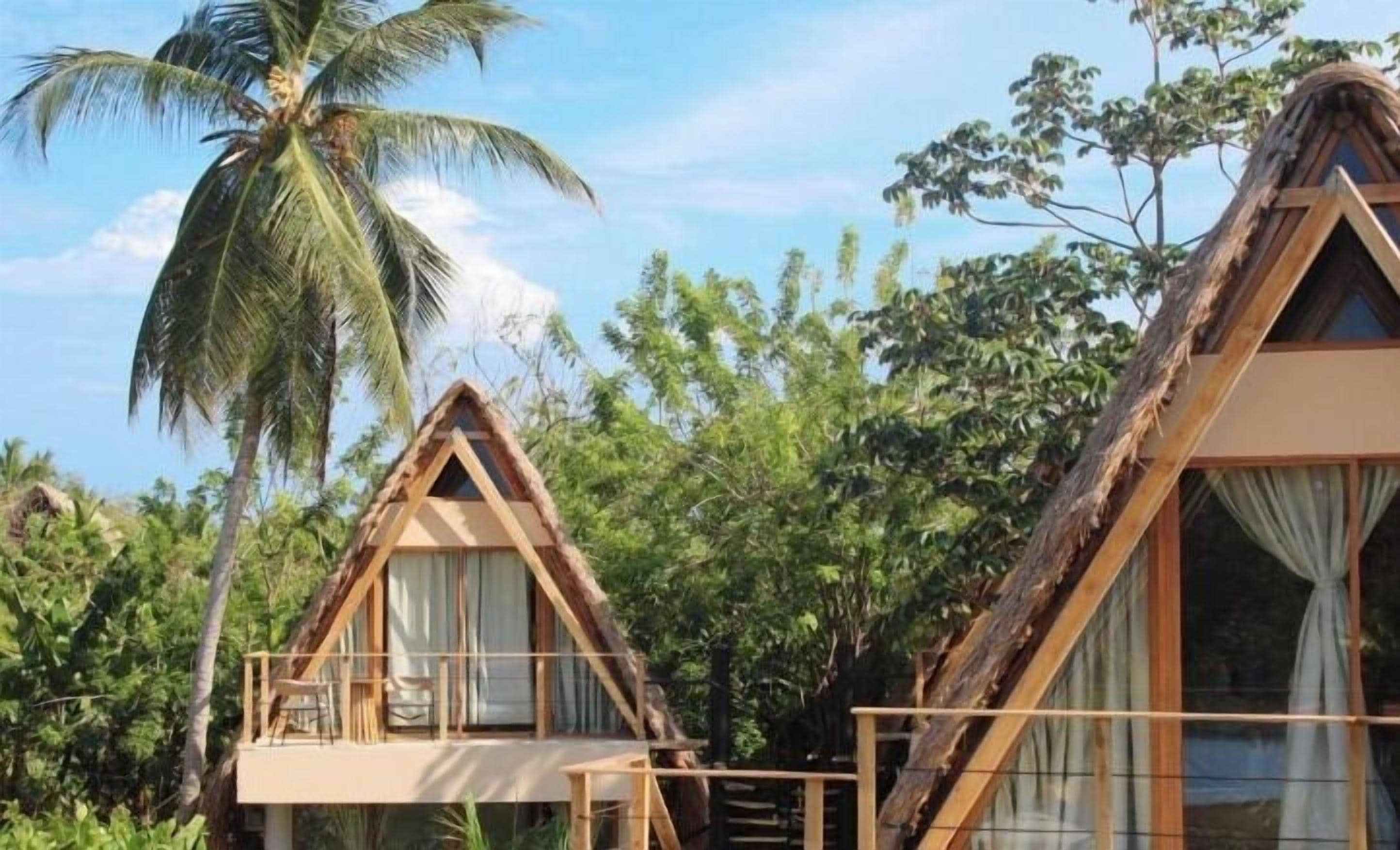 Duas casas de campo com telhados de palha e varandas, rodeadas por vegetação exuberante e uma palmeira.