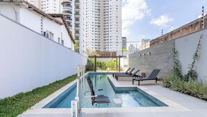 Piscina com espreguiçadeiras, cercada por um muro baixo e um edifício alto ao fundo.