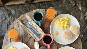Uma mesa de café da manhã com pratos de ovos mexidos, pães, um prato com legumes verdes, xícaras de café e suco.