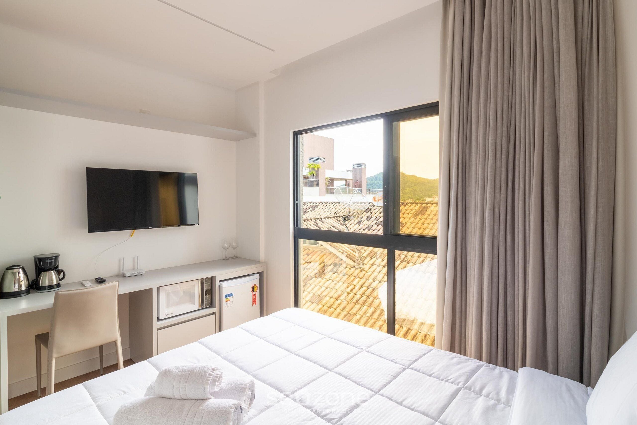 Quarto de hotel com cama, televisão, escrivaninha com cafeteira, micro-ondas e janela com cortinas.