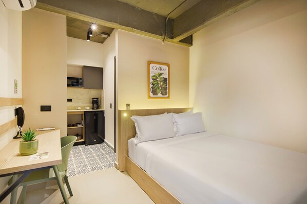 Quarto de hotel pequeno com cama, escrivaninha com planta, micro-ondas e uma pequena cozinha.