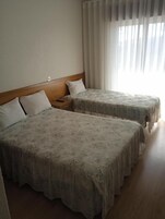 Quarto de hotel com duas camas, cada uma com cabeceira de madeira e colcha florida. Há uma mesinha de cabeceira com abajur e uma tomada na parede.