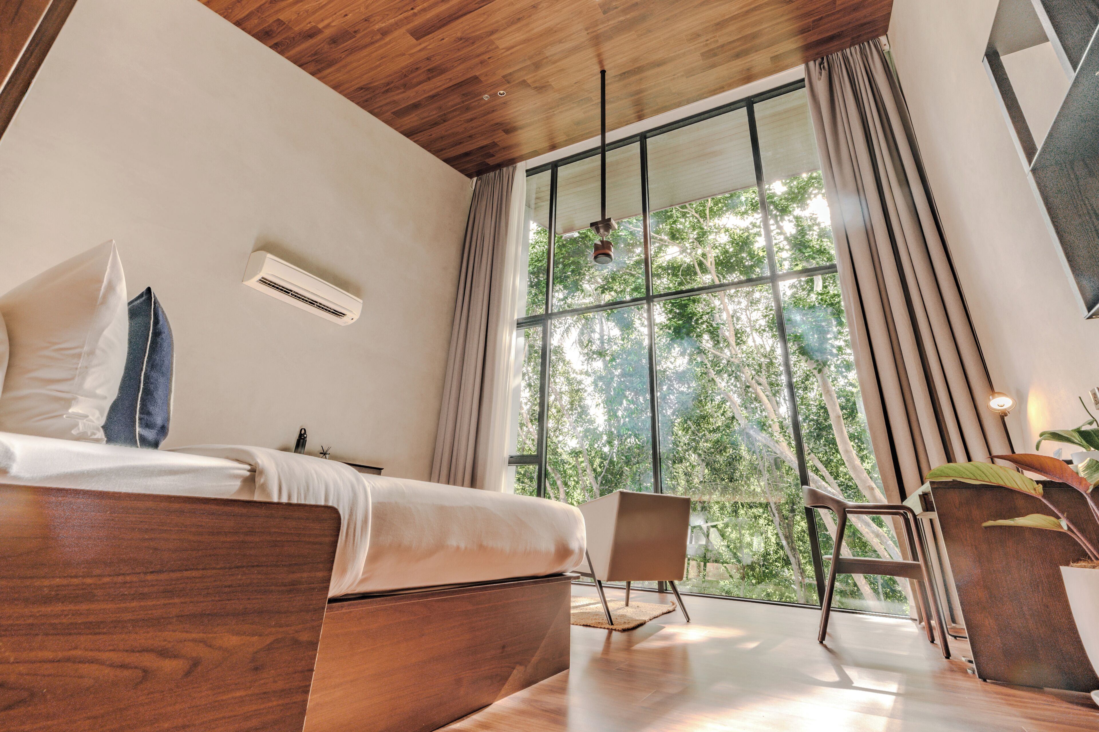 Quarto moderno com teto de madeira, uma janela grande com cortinas, cama, cadeira e escrivaninha.
