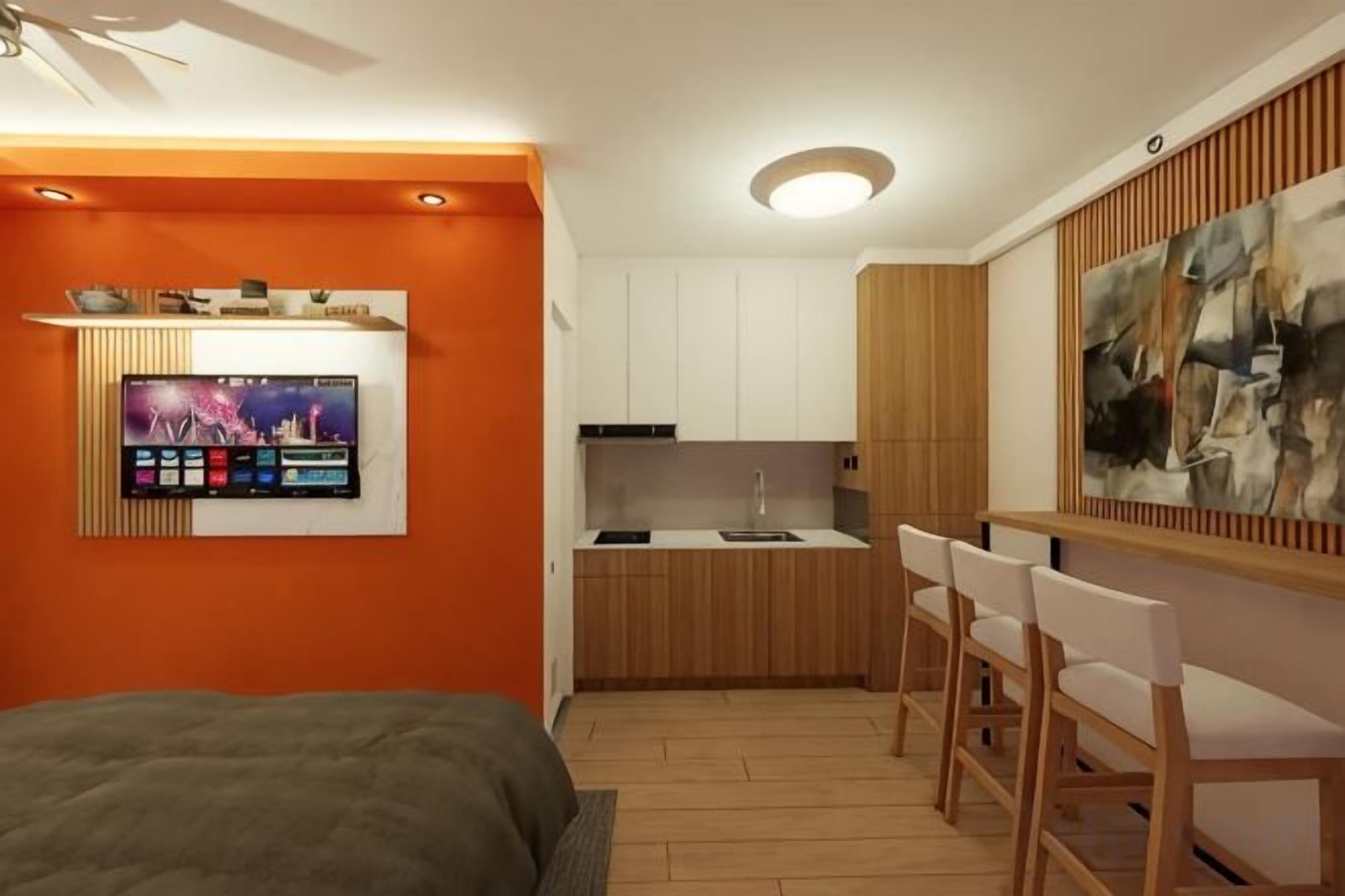 Quarto de hotel compacto com cama, televisão fixada na parede, pequeno espaço de cozinha e área de refeições com cadeiras.