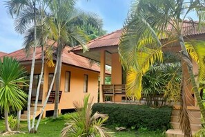 Um edifício com telhado vermelho e várias janelas, cercado por vegetação, incluindo palmeiras e arbustos.