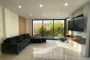 Uma sala de estar moderna com um sofá seccional, uma TV de tela plana e uma grande porta de vidro deslizante que dá para um pátio com plantas.
