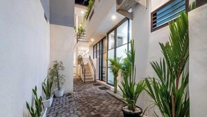 Um corredor estreito e pavimentado, com plantas em vasos, uma escadaria e uma porta de vidro que dá acesso a um andar superior.