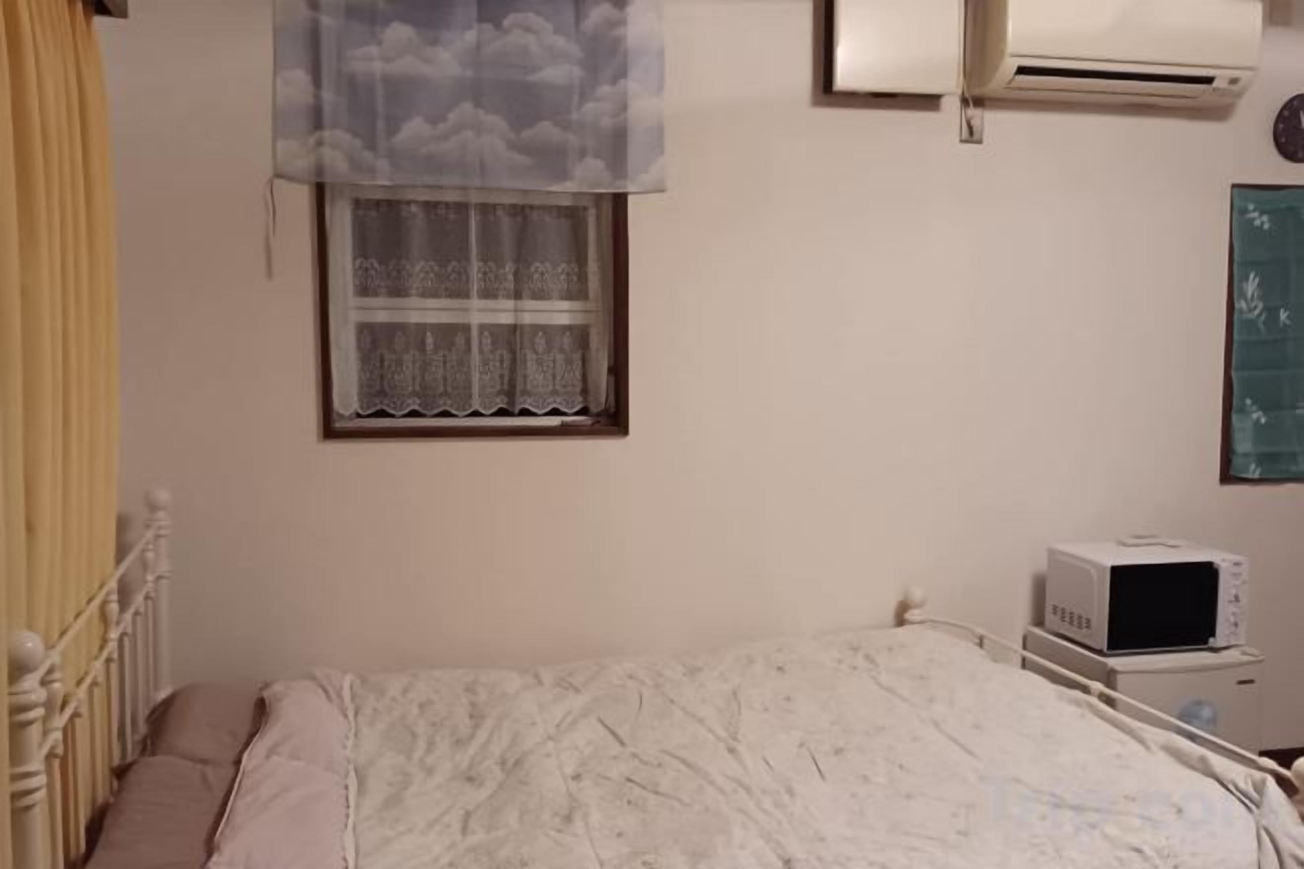 Quarto com cama, janela com cortina de renda, mesa de cabeceira com micro-ondas e ar-condicionado fixado na parede.