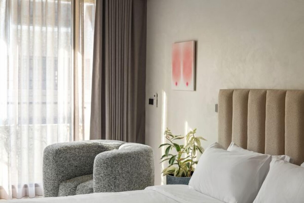 Quarto de hotel com cama, poltrona cinza estofada, planta em vaso e uma obra de arte na parede.