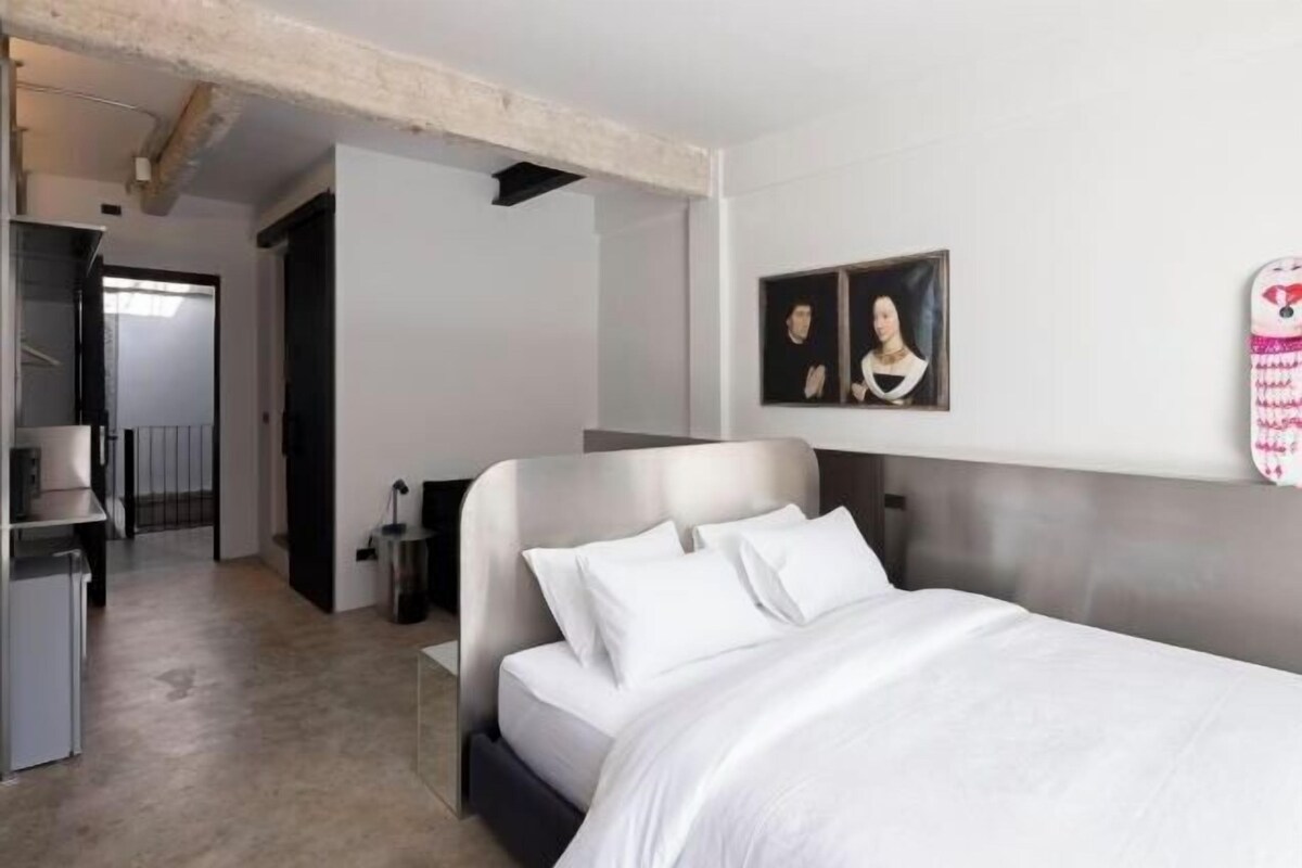 Quarto moderno com uma cama, uma escrivaninha e uma cadeira. Há uma porta que dá para outro quarto.