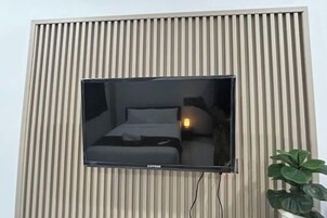 Televisão fixada na parede, com uma cama visível no reflexo.